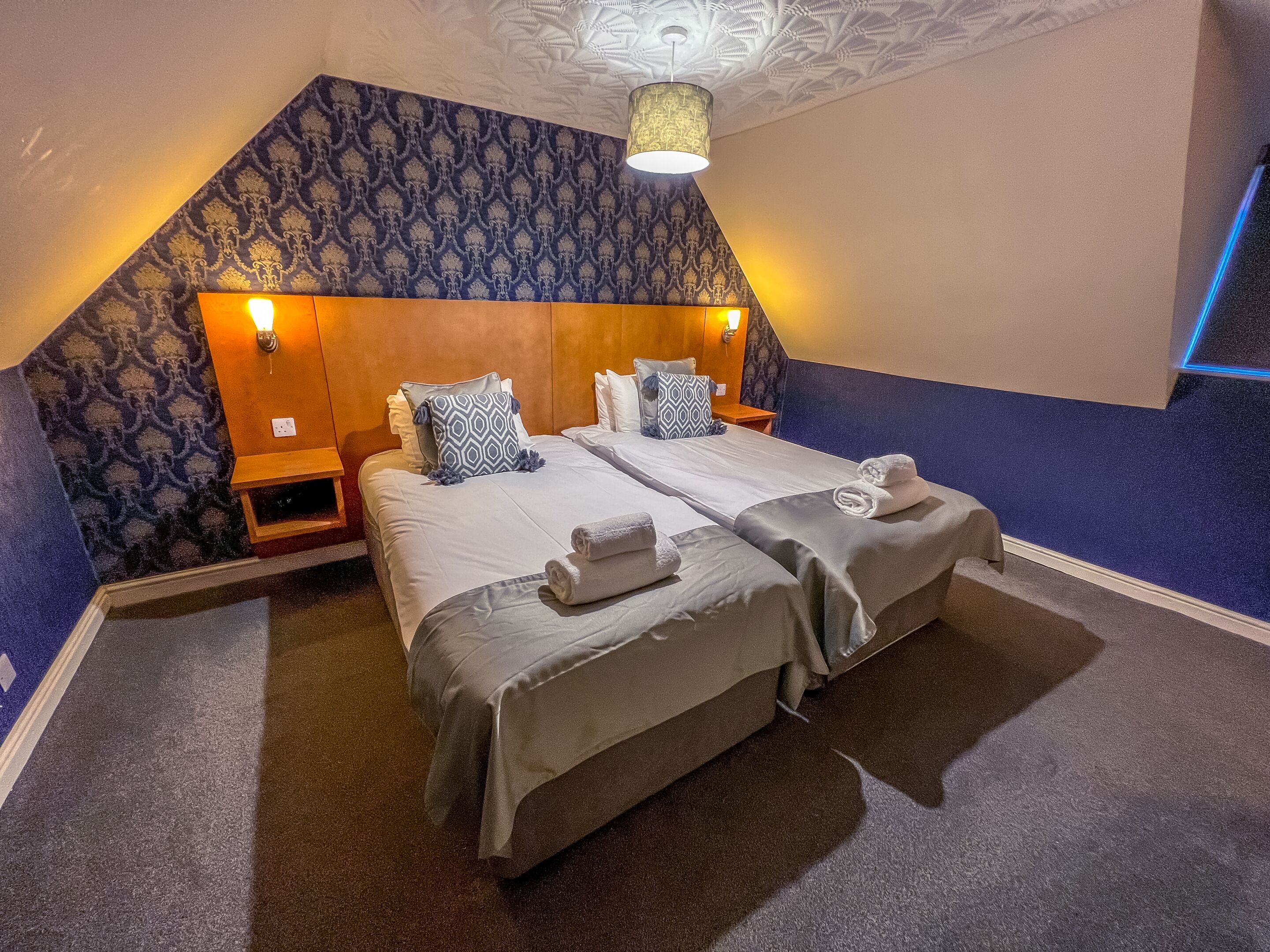 Quarto de hotel com duas camas, papel de parede estampado, cabeceira de madeira e teto com design geométrico.