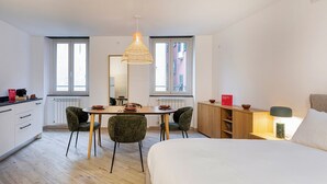 Quarto moderno com uma cama grande, uma mesa de jantar com cadeiras, um lustre e vista para prédios pelas janelas.
