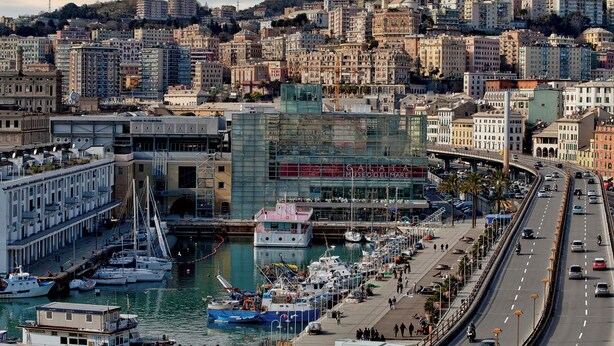 Marina com barcos atracados, estrada com veículos e paisagem urbana ao fundo.