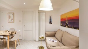 Uma sala de estar moderna com um sofá bege, uma mesa de jantar com cadeiras, um abajur e um quadro com um pôr do sol.
