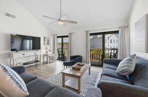 Sala de estar moderna com televisão de tela plana, ventilador de teto, área de jantar com mesa e cadeiras, e varanda com vista para casas.