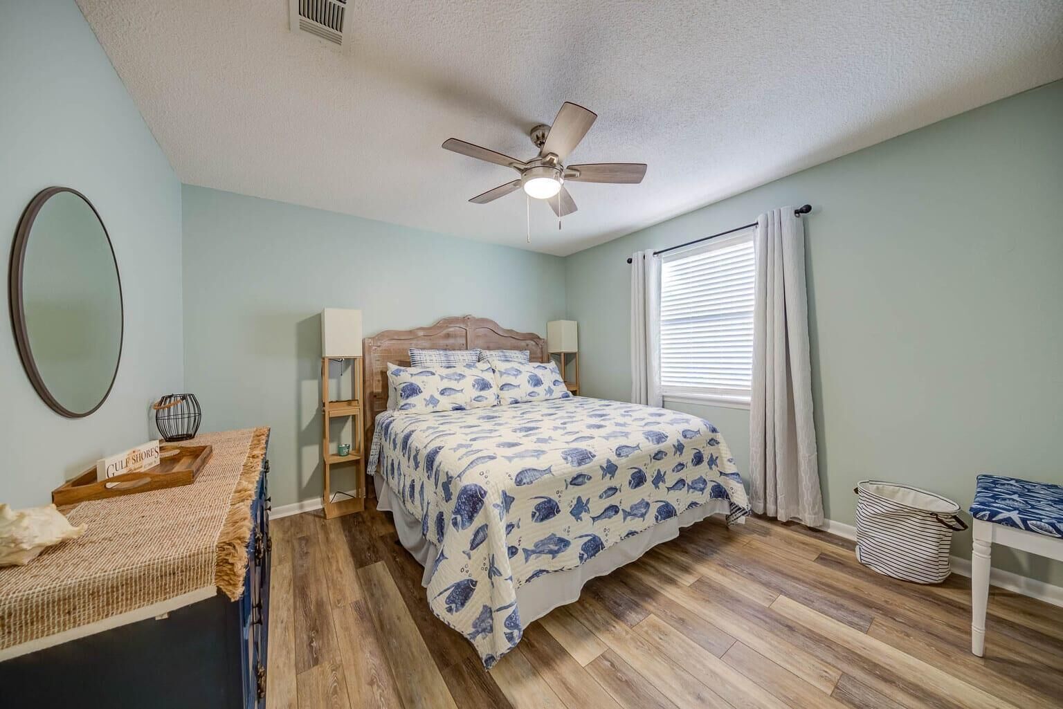 Quarto com cama, ventilador de teto, mesa de cabeceira, abajur, espelho e janela com cortinas.