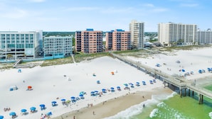 Um hotel à beira-mar com vários edifícios, um cais e uma praia arenosa com guarda-sóis e pessoas.