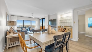 Uma sala de estar moderna com uma janela ampla, uma área de jantar com mesa de madeira e uma cozinha com televisão.