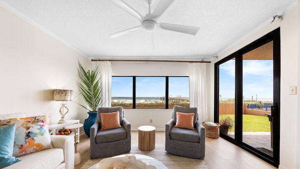 Uma sala de estar espaçosa com ventoinha no teto, uma grande janela com vista para o exterior, um sofá branco com almofadas coloridas, duas poltronas cinzentas e uma mesa de centro redonda com objetos decorativos.