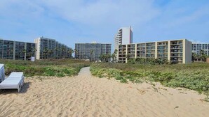 Um hotel à beira-mar com vários edifícios altos, um caminho de areia que leva à praia e pilhas de móveis brancos.