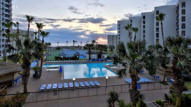 Área de piscina de hotel com uma piscina grande, espreguiçadeiras e palmeiras.