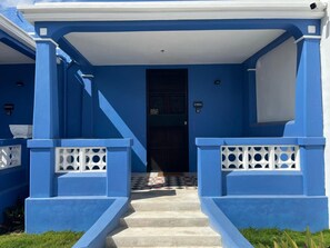 Um edifício azul com varanda branca, porta preta e escadaria que leva à entrada.