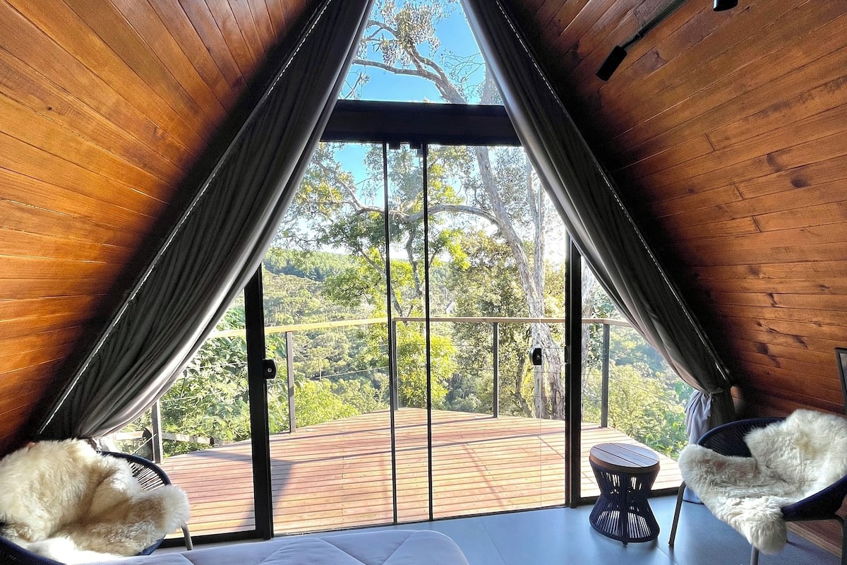 Um quarto acolhedor com teto inclinado, uma cama grande, duas cadeiras e vista para um deck de madeira e árvores.