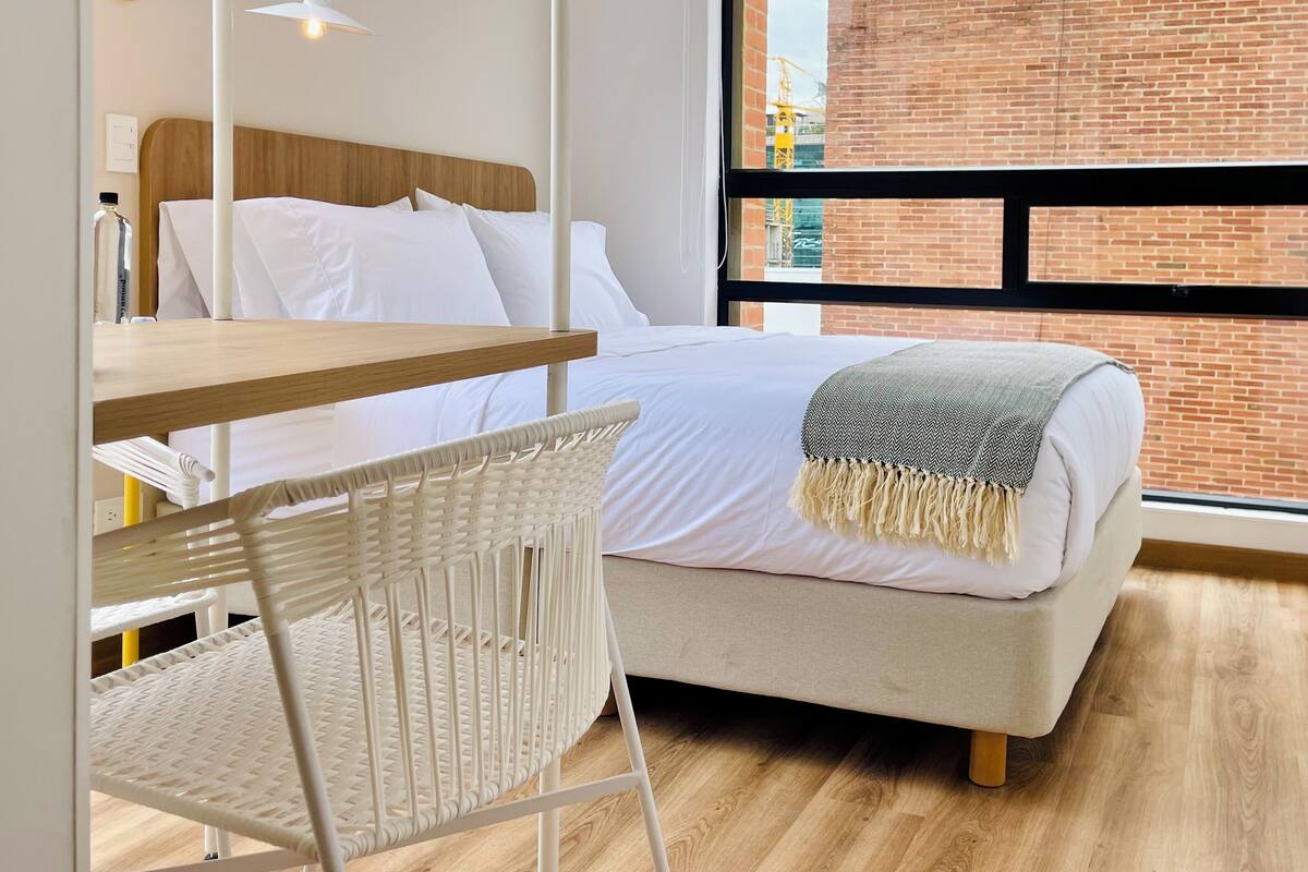 Quarto de hotel moderno com uma escrivaninha de madeira, uma cadeira de vime branca, uma cama com manta cinzenta e uma janela grande com vista para um edifício de tijolos.