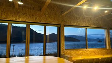 Quarto com grandes janelas que oferecem vista para um lago e montanhas. Há uma mesa de madeira em primeiro plano.