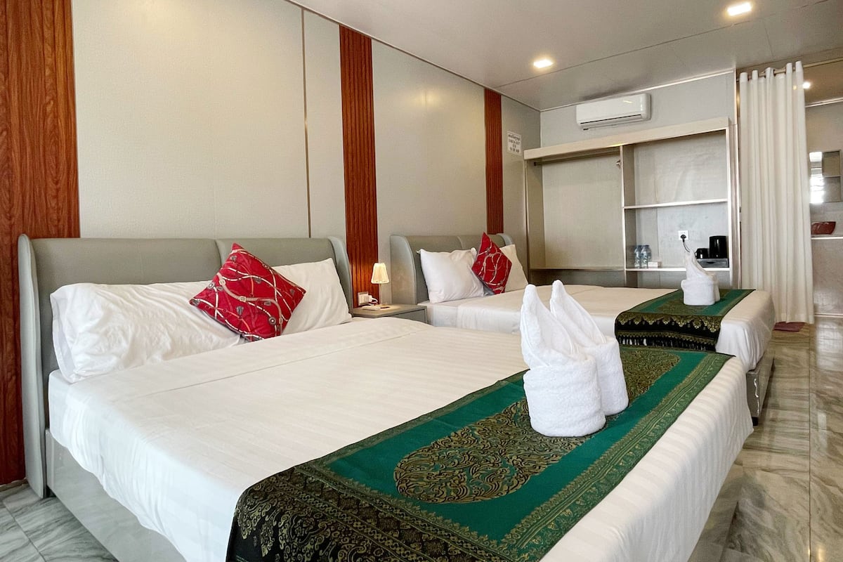 Quarto de hotel com duas camas, cada uma com lençóis brancos e almofadas decorativas vermelhas. Há um corredor com padrão verde e dourado sobre as camas, e uma toalha branca em cada cama. O quarto tem uma parede de destaque em madeira, teto branco e uma prateleira embutida com um espelho e uma cafeteira.