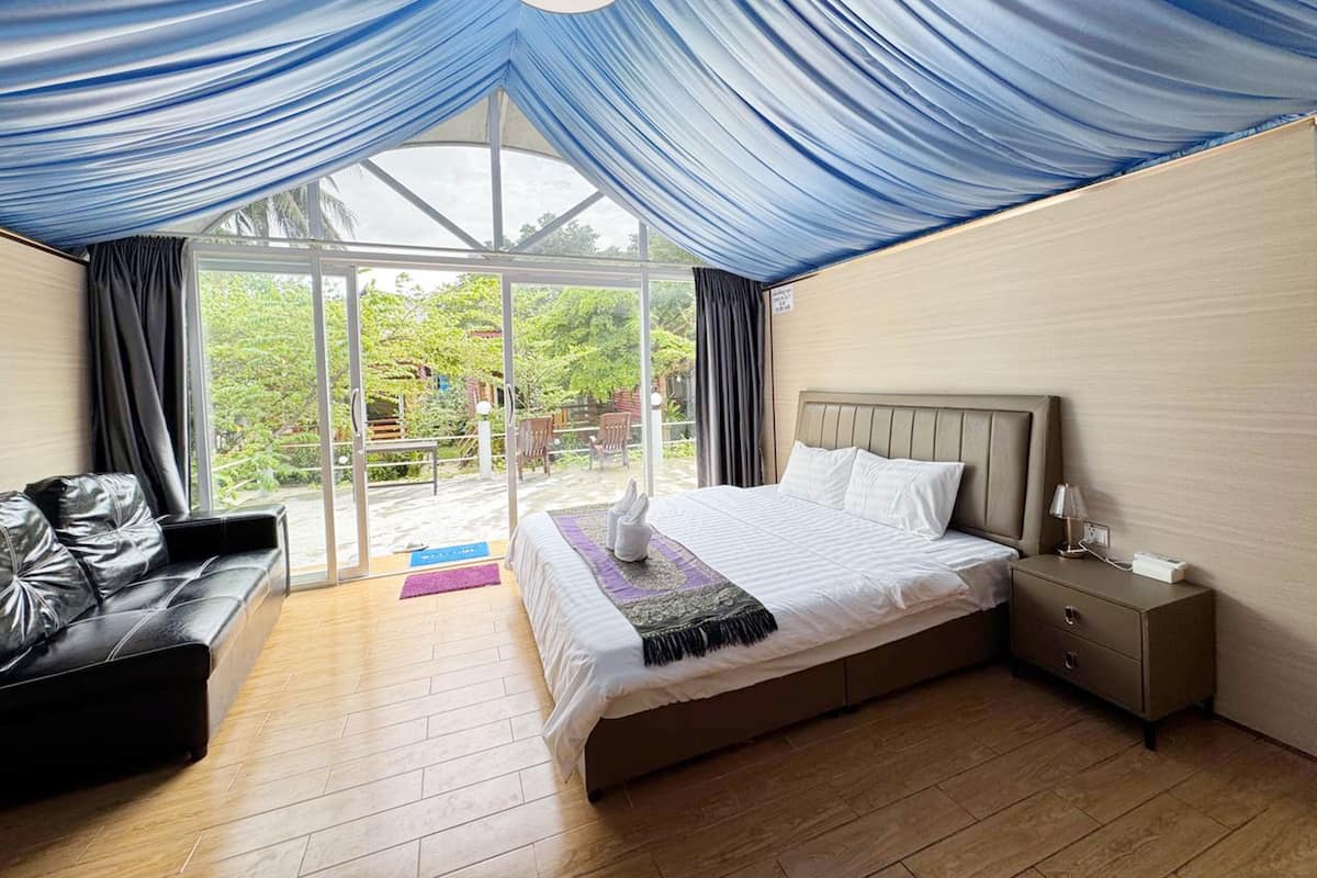 Quarto com cama, mesa de cabeceira, sofá e vista para o verde lá fora.