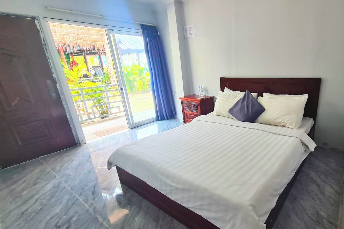 Quarto de hotel com uma cama grande, uma mesa de cabeceira de madeira e vista para um jardim através de uma porta de correr de vidro.