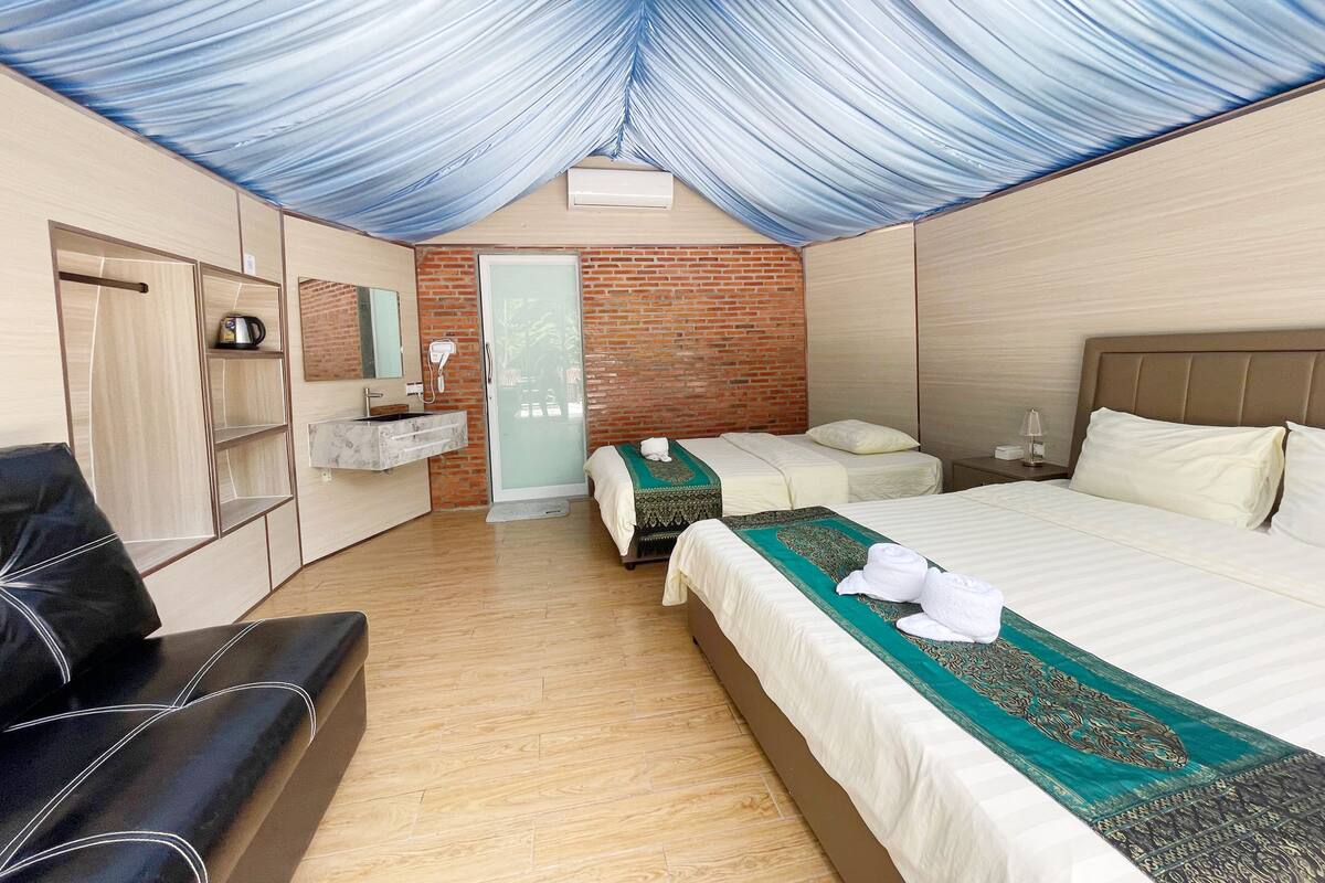 Quarto de hotel com parede de tijolos, porta deslizante, cama com colcha verde e branca, mesa de cabeceira com abajur e sofá de couro preto.
