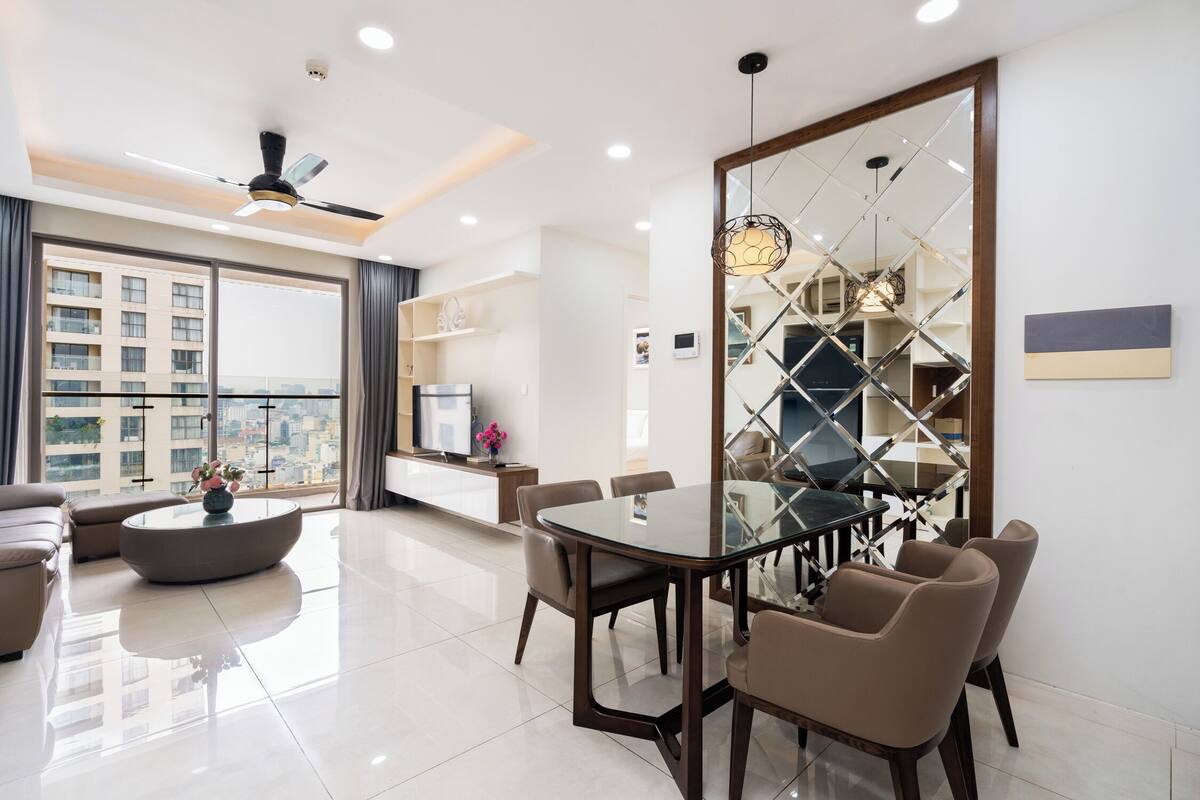 Uma sala de estar moderna com mesa de jantar de vidro, assentos confortáveis, ventilador de teto e uma grande janela com vista para a cidade.