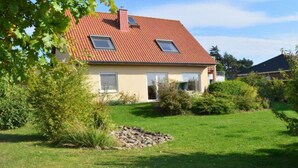 Uma casa térrea com telhado de telhas vermelhas, um pequeno jardim frontal e uma área de jardim com diversas plantas e uma formação rochosa.