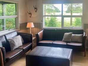 Uma sala de estar com um sofá de couro marrom escuro, um banquinho combinado e uma luminária. Há duas janelas com vista para o verde lá fora.