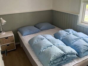 Uma cama de solteiro com roupa de cama e almofadas azuis, uma luminária de cabeceira e uma pequena cesta de vime sobre a mesinha de cabeceira.