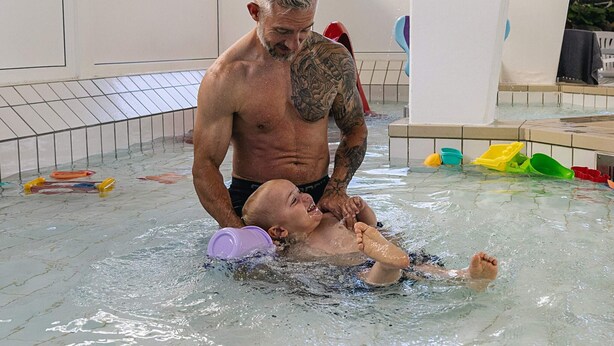 Um homem em uma piscina com uma criança, cercado por diversos brinquedos de piscina.
