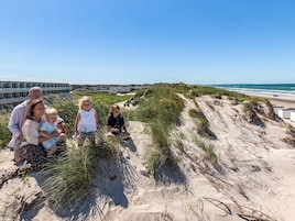 Família em uma praia arenosa com dunas e um edifício ao fundo.