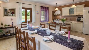 Uma área de jantar com uma mesa posta para quatro pessoas, uma cozinha com armários brancos e um relógio na parede.