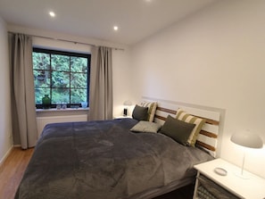 Quarto com uma cama grande, janela com cortinas, mesa de cabeceira com abajur e vista para o verde lá fora.