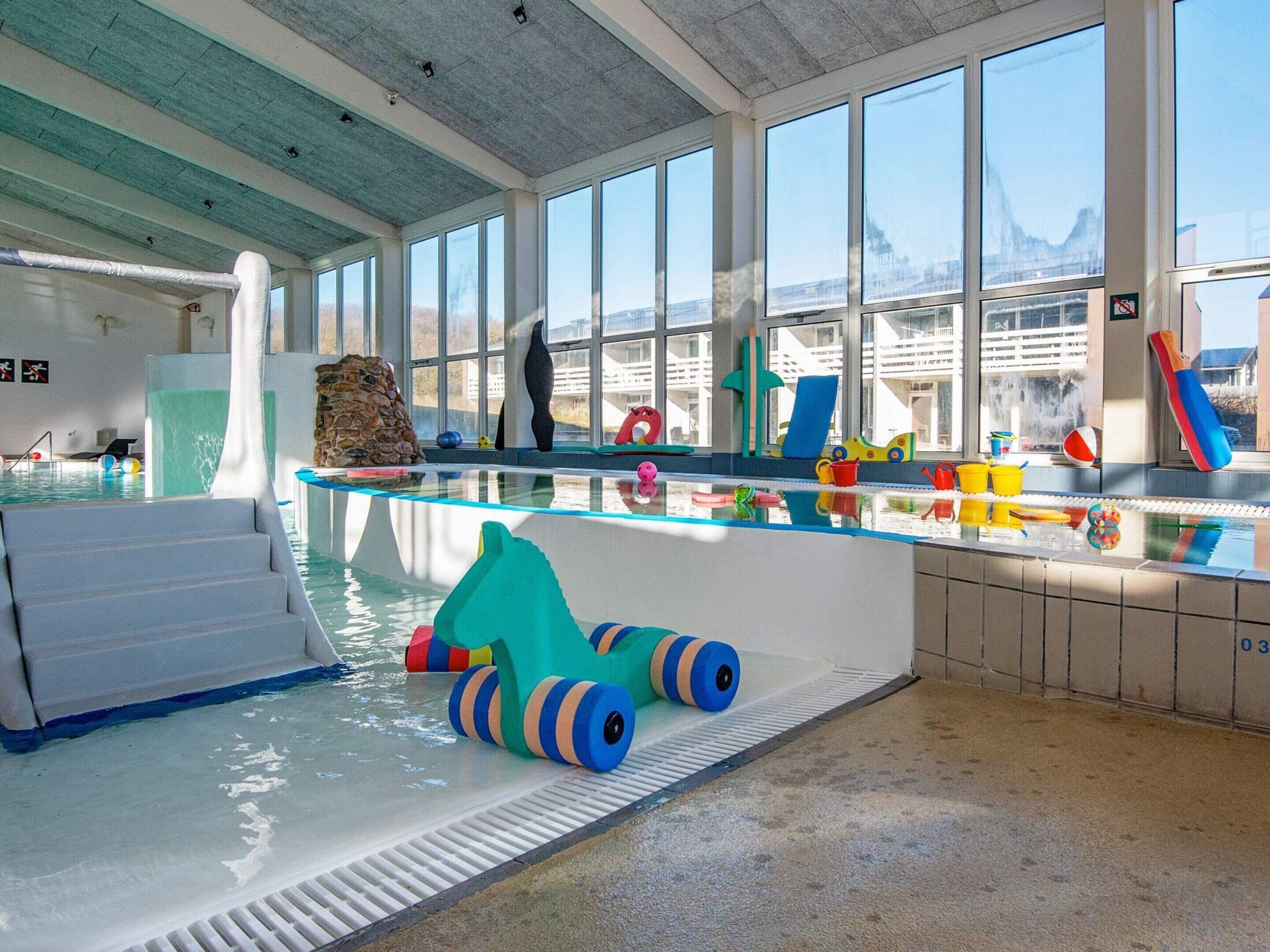 Piscina interior com barreira de vidro, parede de pedra e diversos brinquedos e equipamentos coloridos.