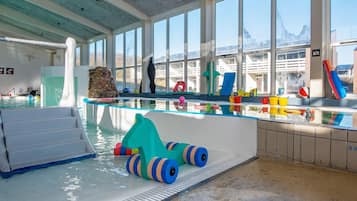 Piscina interior com barreira de vidro, parede de pedra e diversos brinquedos e equipamentos coloridos.