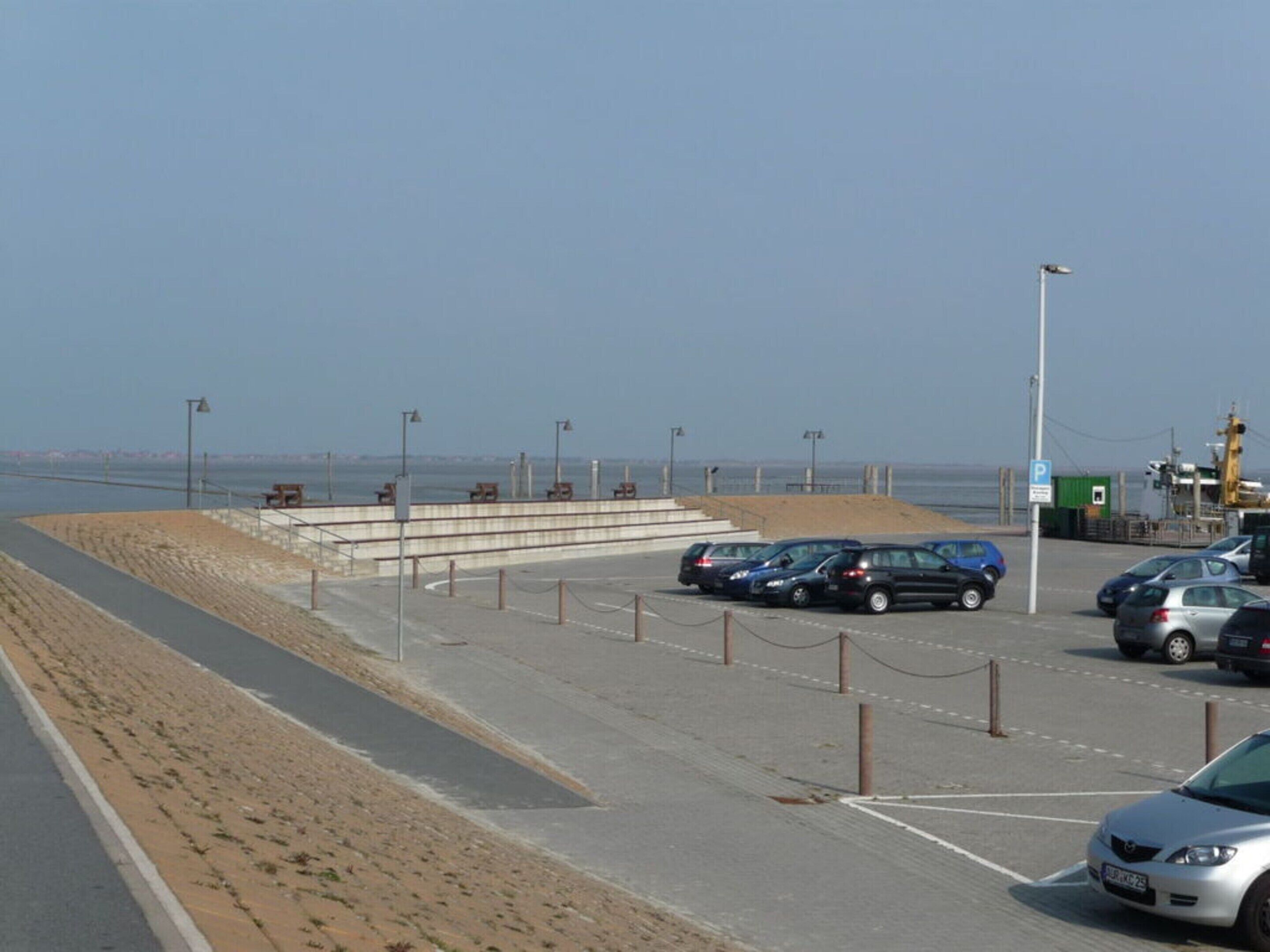 Uma área de estacionamento com carros, um caminho para pedestres e vista para o mar.