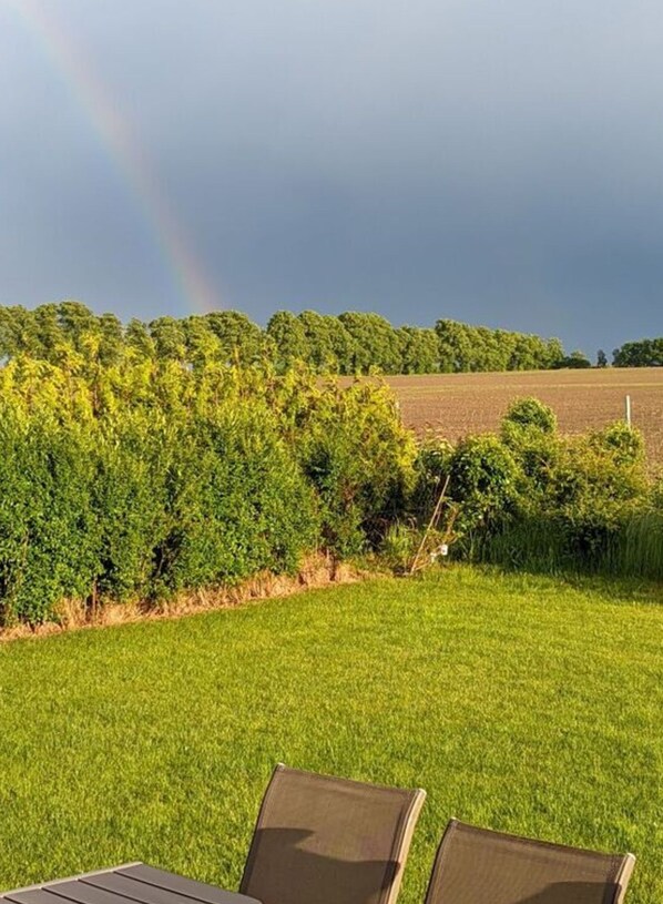 Uma área externa gramada com uma mesa e cadeiras, um arco-íris no céu e um campo ao fundo.