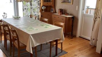 Uma sala de jantar com mesa posta para quatro pessoas, cadeiras de madeira, toalha de mesa estampada, luminária e armário com utensílios de cozinha.