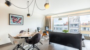 Uma área de refeições moderna com uma mesa posta para quatro pessoas, um sofá preto e uma janela grande com vista para a cidade.