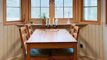 Uma sala de jantar com mesa e cadeiras de madeira, grandes janelas com vista para árvores e um castiçal sobre a mesa.