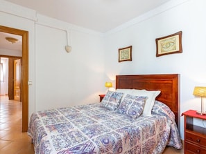 Quarto com cabeceira de madeira, colcha estampada, duas luminárias de apoio e duas imagens emolduradas na parede.