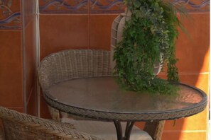 Uma mesa de vidro redonda com uma cadeira de vime ao lado, uma planta pendente e uma parede de azulejos com um padrão decorativo.