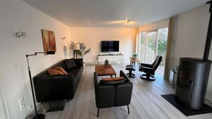 Uma sala de estar moderna com sofá, poltrona e banquinho, TV, lareira e uma janela grande.