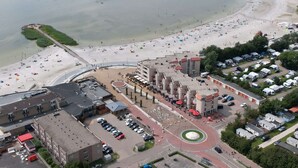 Vista aérea de uma área costeira com praia, parque de estacionamento e um complexo de edifícios com vários andares e um pátio central.