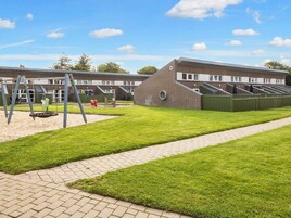 Um parquinho com balanços e um caixa de areia, um edifício de tijolos com telhado inclinado e um caminho pavimentado.