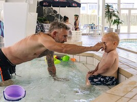 Um homem e uma criança numa piscina, com um muro de pedra e guarda-sóis ao fundo.