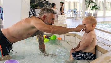 Um homem e uma criança numa piscina, com um muro de pedra e guarda-sóis ao fundo.