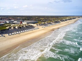 Uma praia com uma fileira de cabanas de praia brancas, um edifício grande com várias janelas e uma faixa de areia com ondas.