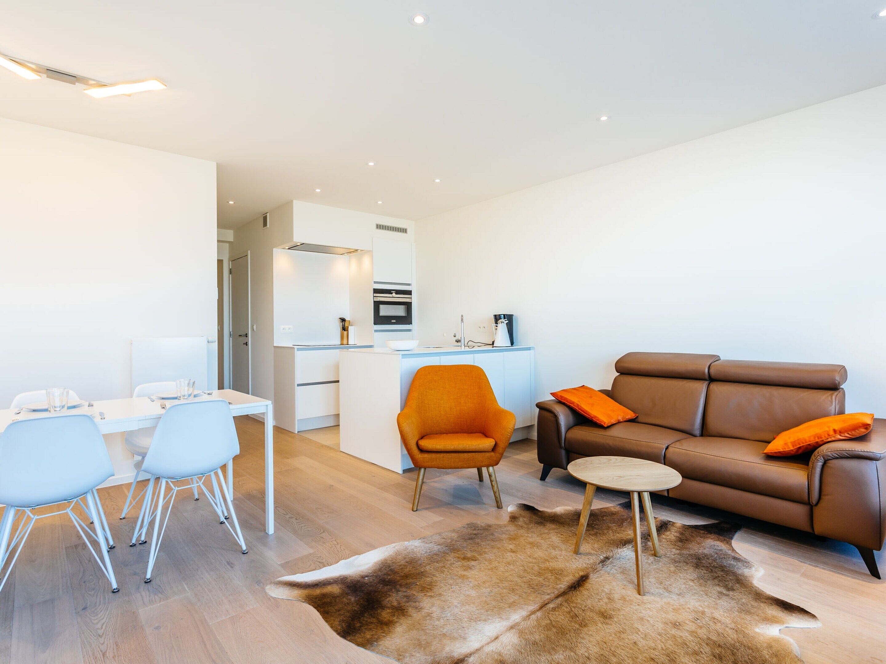Uma sala de estar moderna com sofá de couro marrom, uma mesinha redonda e um espaço de refeições com mesa branca e cadeiras azuis.