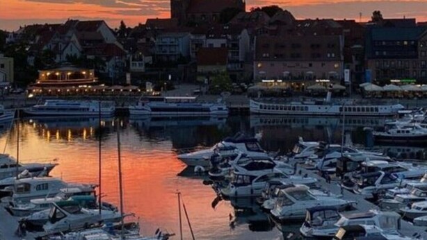 Marina com barcos atracados, um edifício notável com torre e céu ao pôr do sol.
