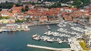 Uma marina com vários barcos atracados, rodeada por uma cidade com edifícios de telhados vermelhos e uma torre de igreja imponente.
