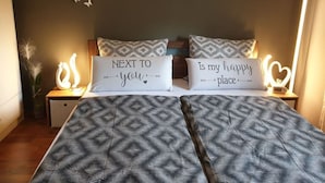 Cama bem arrumada com roupa de cama estampada, dois travesseiros com texto, uma arte na parede de uma praia rochosa e um objeto decorativo na parede.