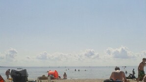 Uma praia com pessoas nadando e tomando sol, céu claro e barcos ao longe.