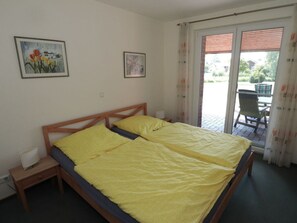 Quarto individual com uma cama e cabeceira de madeira, colcha amarela, mesa de cabeceira pequena com abajur e janela com cortinas.