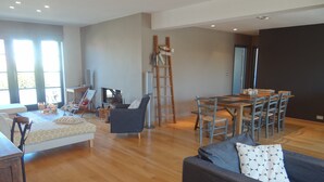 Uma sala de estar ampla com piso de madeira, um sofá cinza escuro, uma mesa de jantar com cadeiras, uma TV e uma escada encostada na parede.