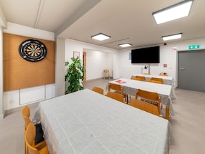 Uma sala de conferências com mesas, cadeiras, um alvo de dardos, uma planta e uma televisão na parede.